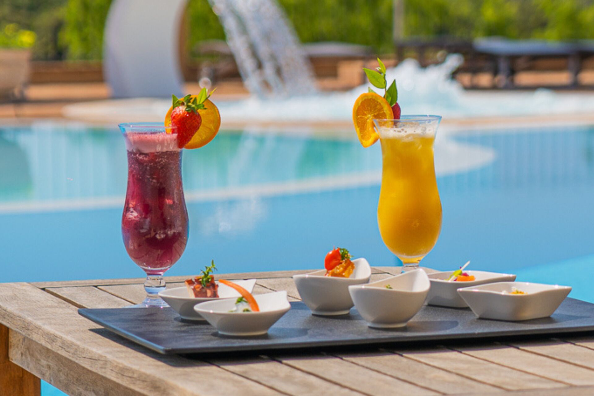 Dois cocktails com guarnições de frutas sobre uma mesa de madeira junto a uma piscina com um toboágua ao fundo.