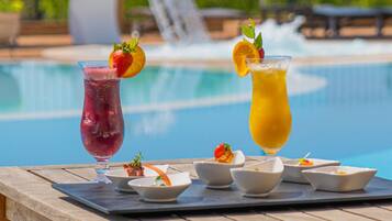 Dois cocktails com guarnições de frutas sobre uma mesa de madeira junto a uma piscina com um toboágua ao fundo.