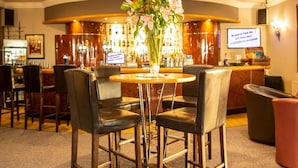Uma área de bar com uma mesa redonda e quatro cadeiras, um vaso grande com flores e um balc ão de bar com garrafas.