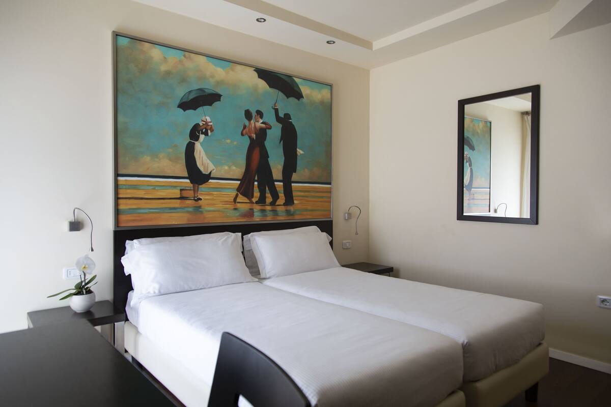 Quarto de hotel com uma cama grande, uma mesa de cabeceira com uma planta, um quadro de um casal numa praia e um espelho na parede.