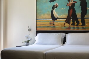 Quarto de hotel com uma cama, um quadro de um casal dançando, uma planta em vaso e uma mesa de cabeceira.