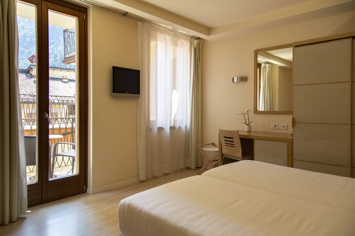Quarto de hotel com cama, escrivaninha, cadeira, televisão, varanda com uma cadeira e porta de correr.