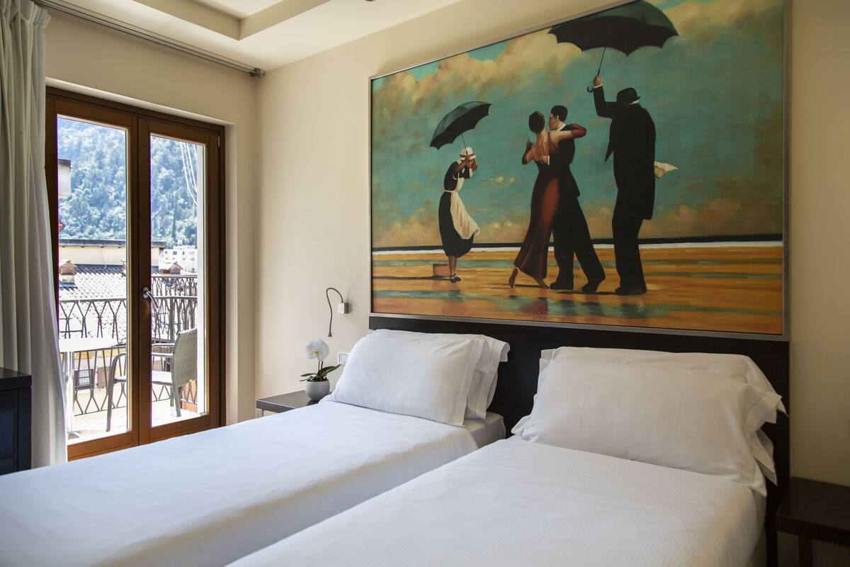 Quarto de hotel com duas camas, um quadro de um casal numa varanda e vista para a área da varanda exterior.