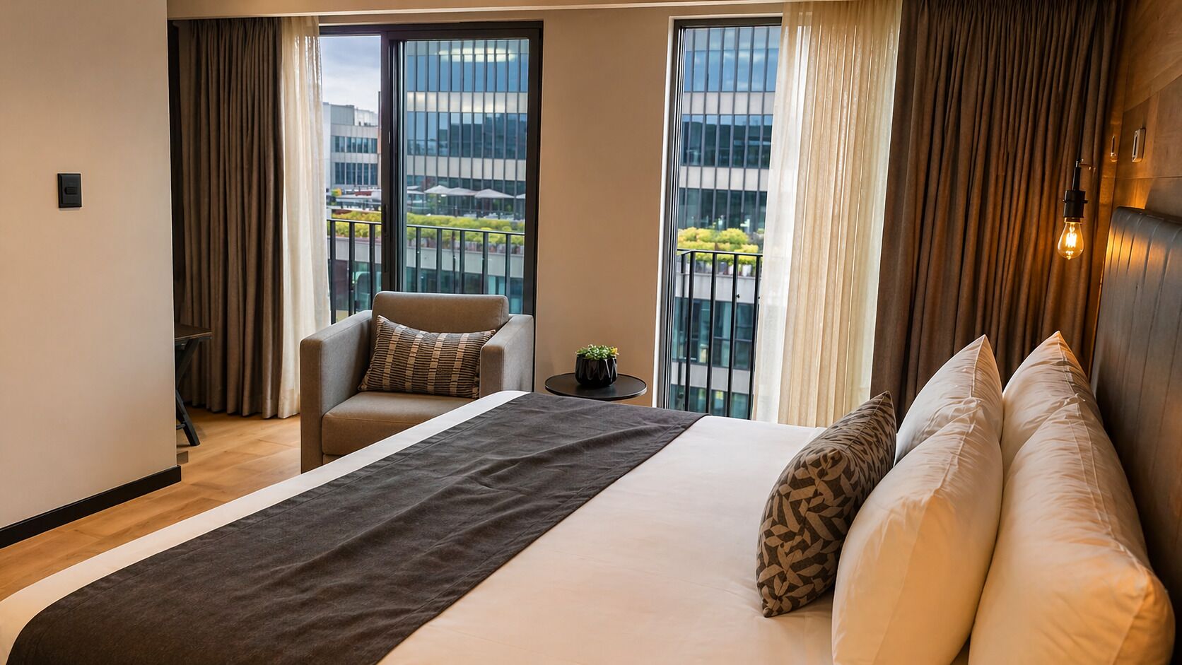 Quarto de hotel com uma cama grande, uma cadeira, uma mesinha com uma planta e vista para edifícios pelas janelas.