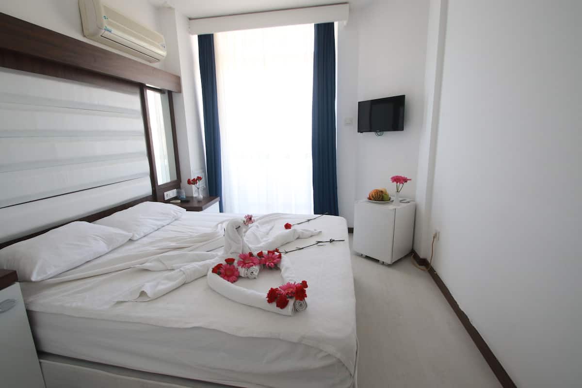 Cama bem arrumada com lençóis brancos e flores rosa, mesa de cabeceira com um vaso de flores e uma TV de tela plana fixada na parede.