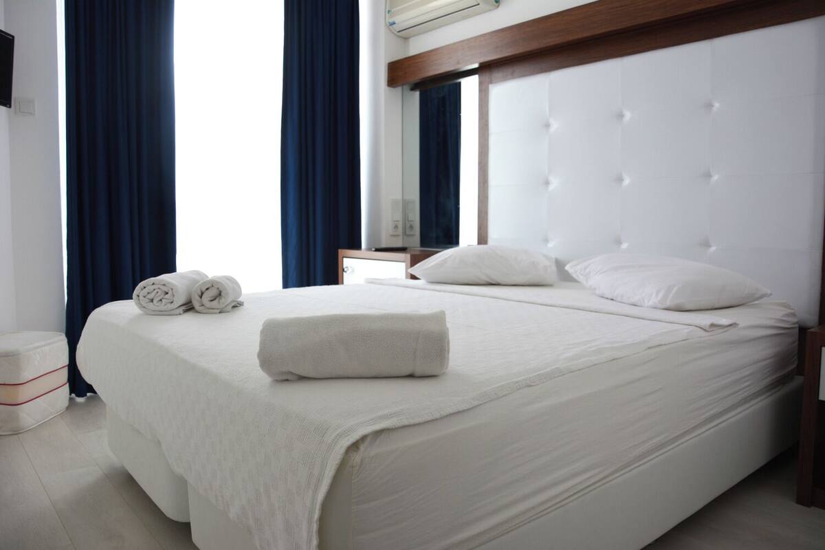 Uma cama bem arrumada com lençóis e almofadas brancas, uma mesa de cabeceira com abajur e uma janela com cortinas azuis.