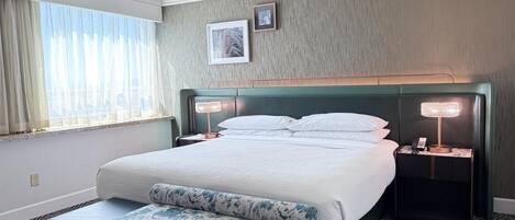 Quarto de hotel com cama, luminárias de cabeceira, um banco, janela com cortinas e um quadro na parede.