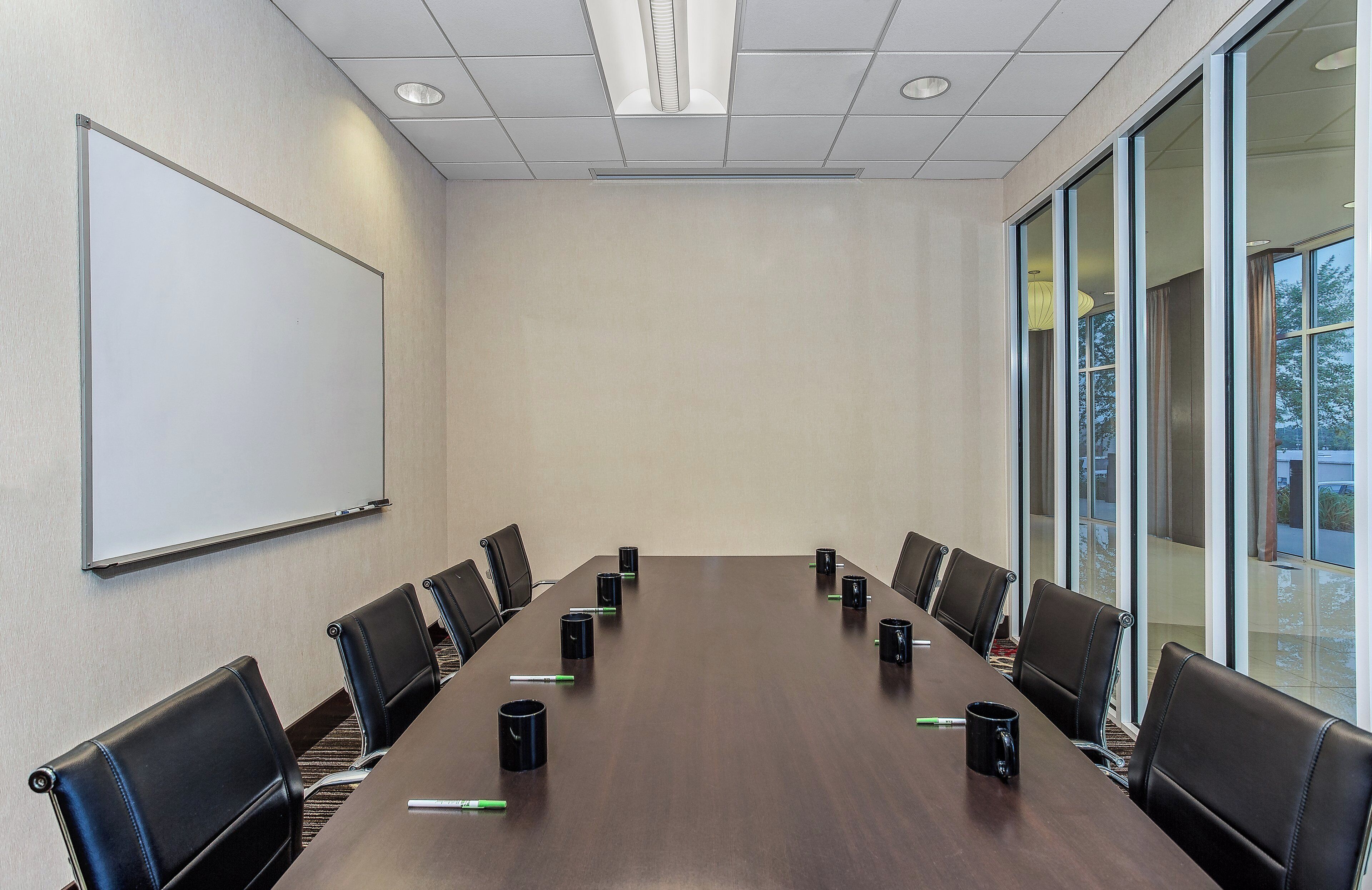 Uma sala de conferências com uma mesa comprida, cadeiras pretas, um quadro branco e uma grande janela com vista para árvores.