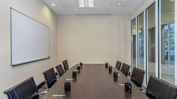 Uma sala de conferências com uma mesa comprida, cadeiras pretas, um quadro branco e uma grande janela com vista para árvores.
