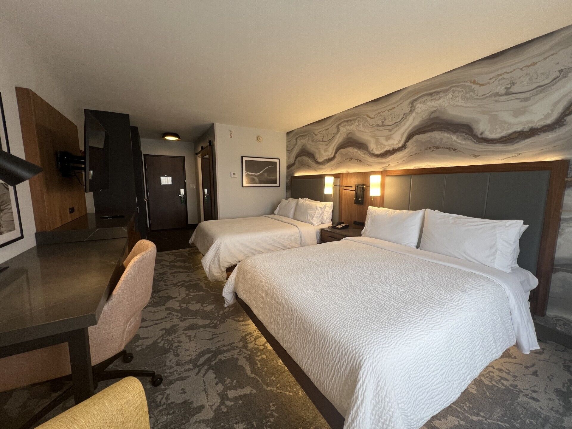 Quarto de hotel com duas camas, uma escrivaninha, uma cadeira e teto com padrão marmorizado.