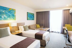 Quarto de hotel com duas camas, um sofá, uma escrivaninha e uma cadeira. Há quadros nas paredes e um aparelho de ar condicionado.