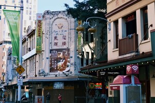 Uma rua com vários edifícios, incluindo um hotel chamado 'COVENT GARDEN HOTEL' e um restaurante chamado 'EMERSON'S GARDEN'. Há também placas para 'Sydney Science Festival' e 'Telstra'.