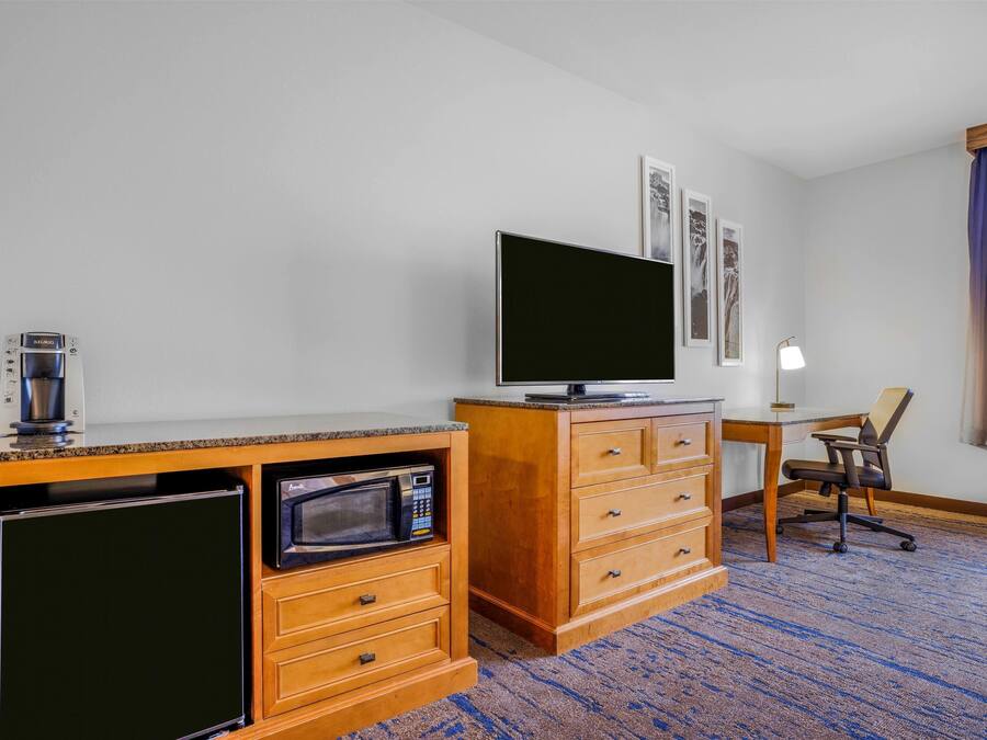 Quarto de hotel com escrivaninha de madeira, televisão, micro-ondas, máquina de café e uma cadeira.