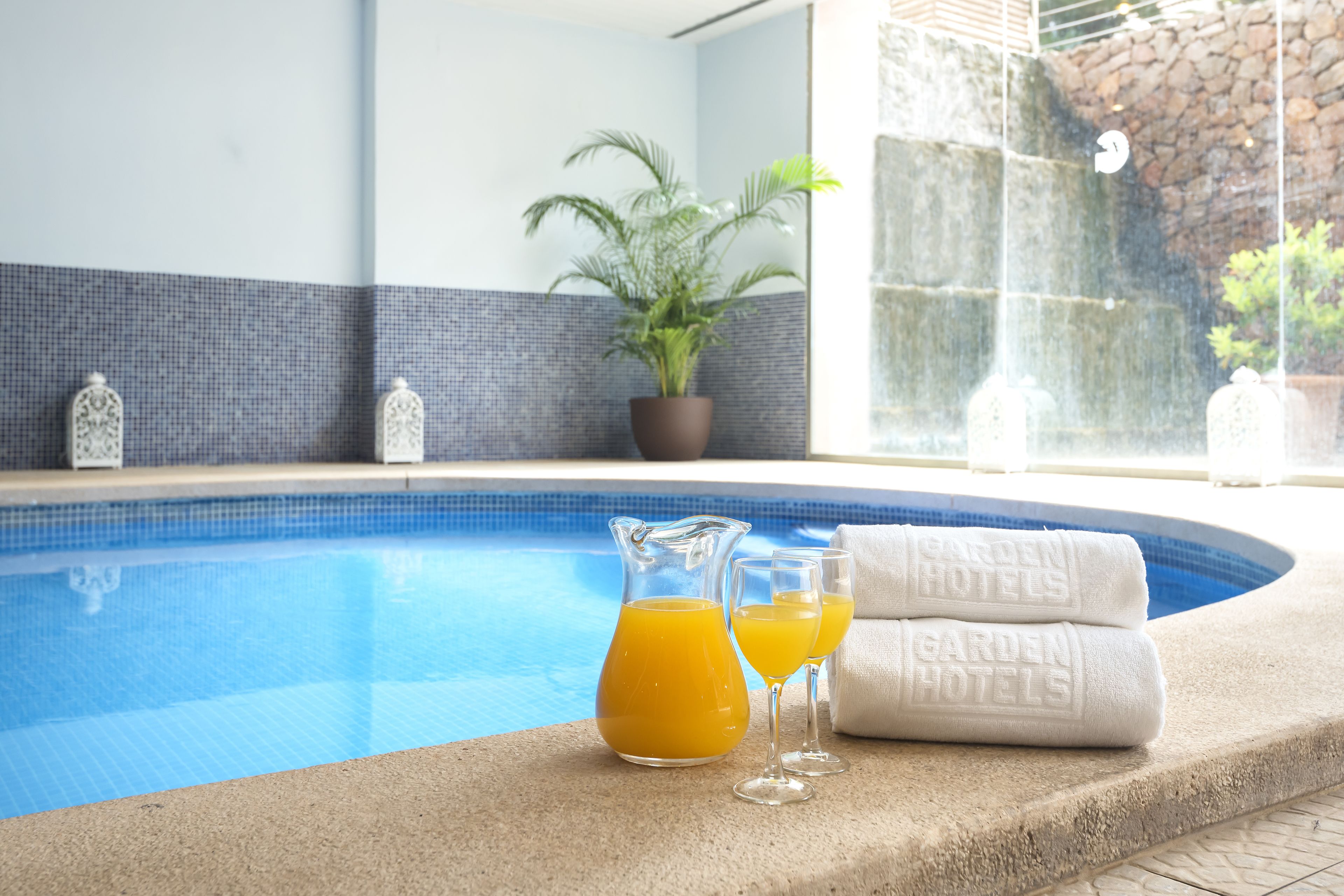 Uma área à beira da piscina com piscina azul cristalina, um vaso de planta e toalhas com a marca 'Garden Hotels'.