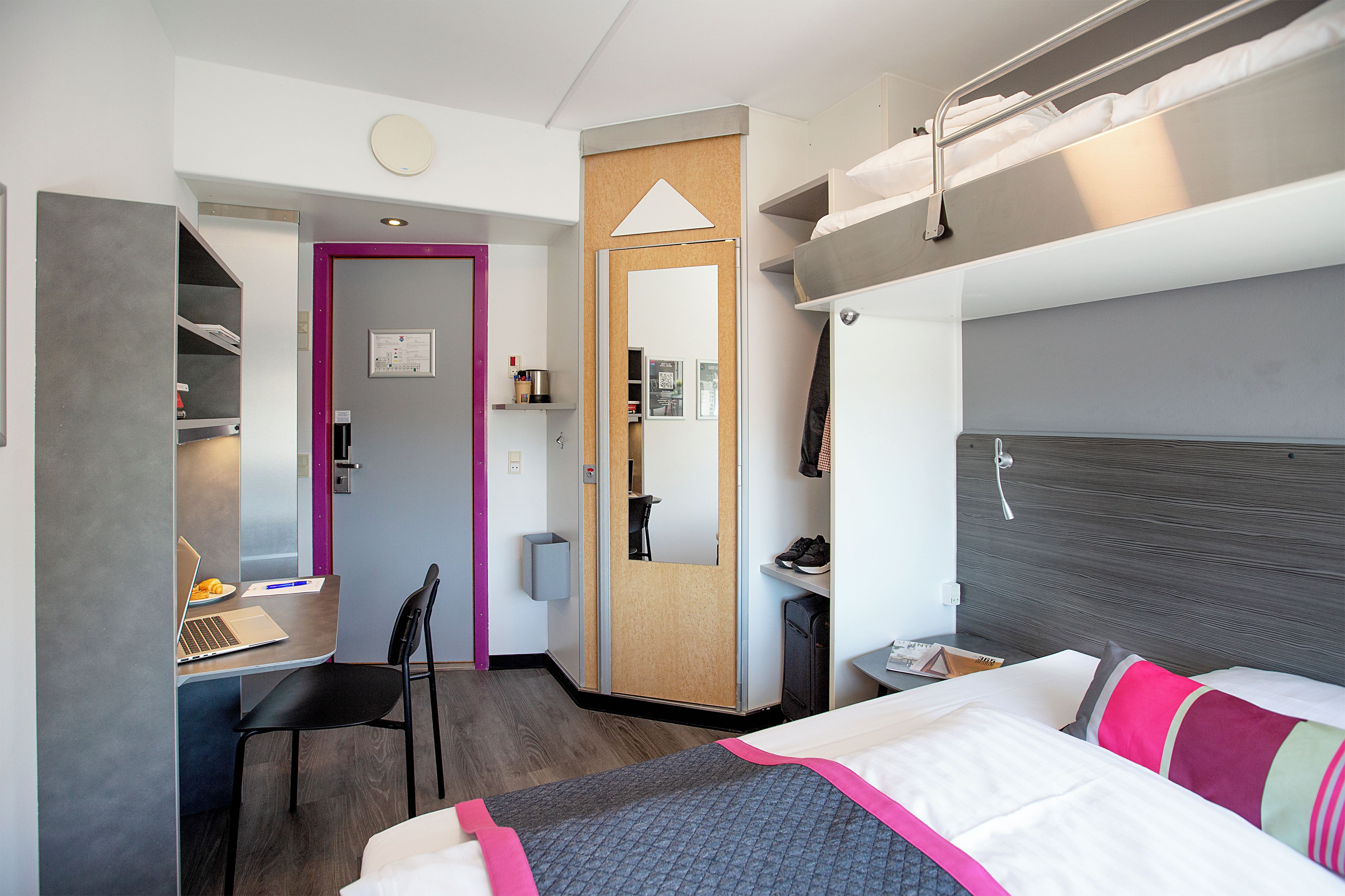 Quarto de hotel com cama, escrivaninha, laptop, cadeira, guarda-roupa e espelho.