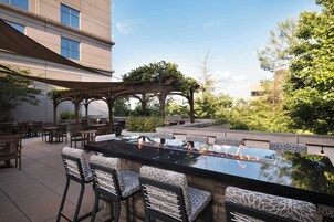 Uma área de bar ao ar livre com uma mesa comprida e várias cadeiras, cercada por vegetação e uma pérgola.