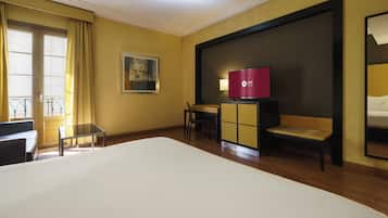 Quarto de hotel com uma cama grande, uma escrivaninha com cadeira, televisão e um quadro na parede.