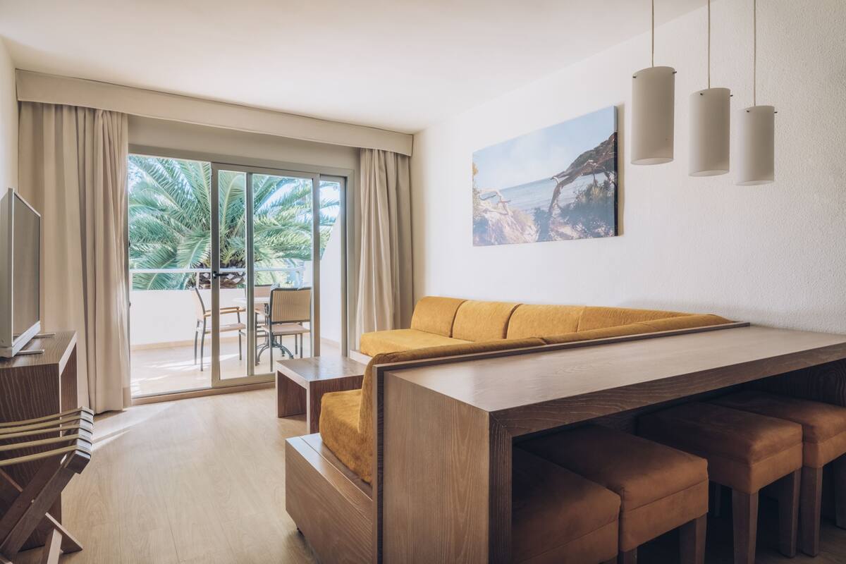Quarto de hotel moderno com uma mesa de jantar de madeira, um sofá e uma janela grande com vista para palmeiras e uma montanha.