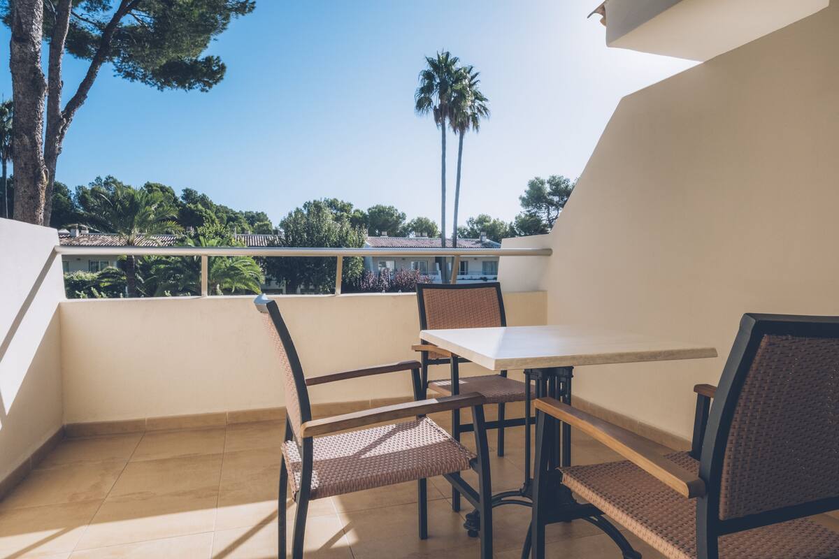 Uma varanda com mesa e cadeiras, de frente para uma área residencial com árvores e céu claro.
