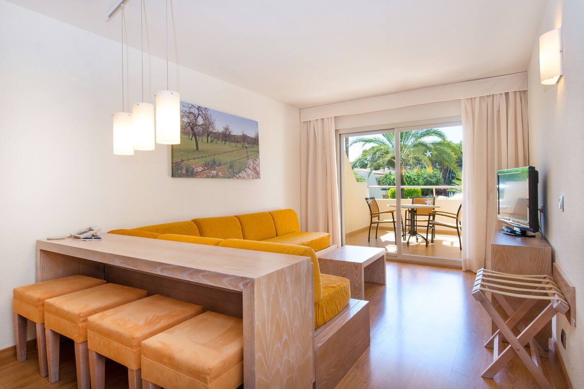 Uma sala de estar moderna com um sofá, uma mesa de centro e uma televisão. Há uma varanda com vista para palmeiras.