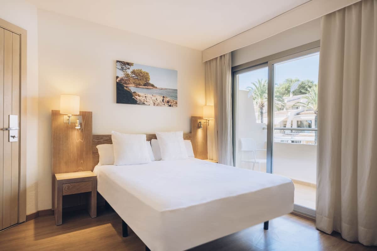 Quarto de hotel com uma cama grande, duas mesas de cabeceira com luminárias, um quadro na parede e vista para o exterior através de uma grande janela.