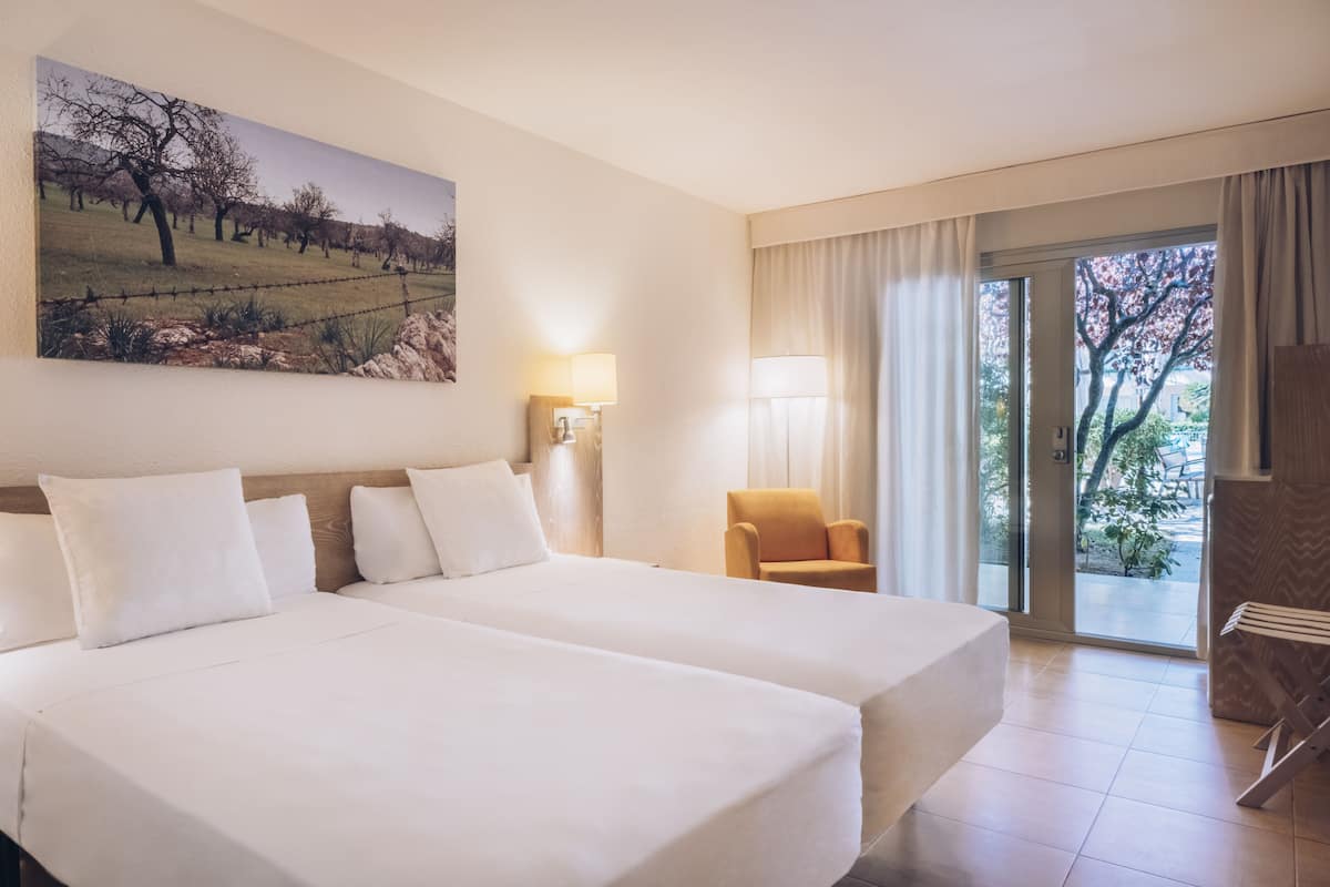 Quarto de hotel com duas camas, uma cadeira, uma luminária e uma janela grande com cortinas.