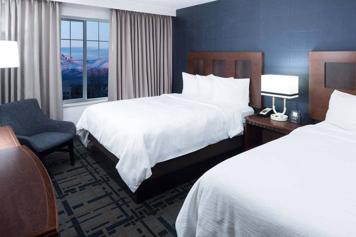 Quarto de hotel com duas camas, uma escrivaninha, uma cadeira, mesa de cabeceira com abajur e vista para uma paisagem nevada pela janela.