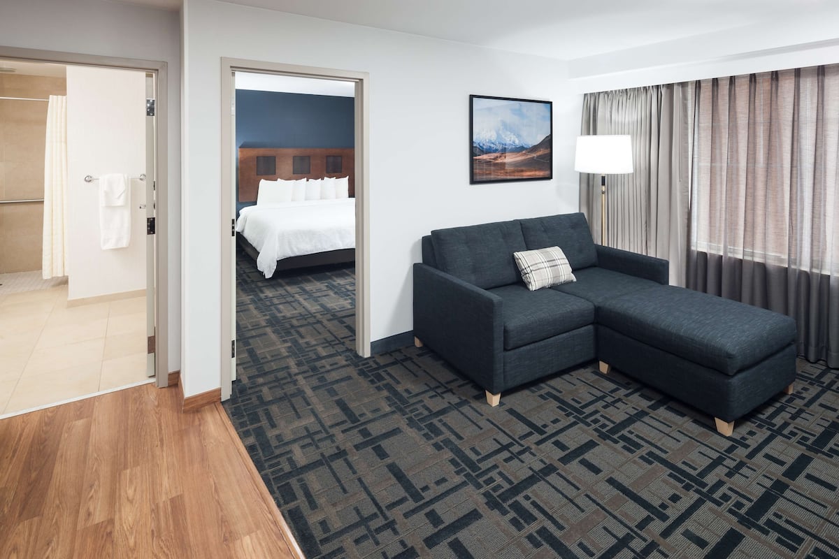 Quarto de hotel com cama, sofá escuro, tapete estampado, quadro na parede e janela com cortinas.
