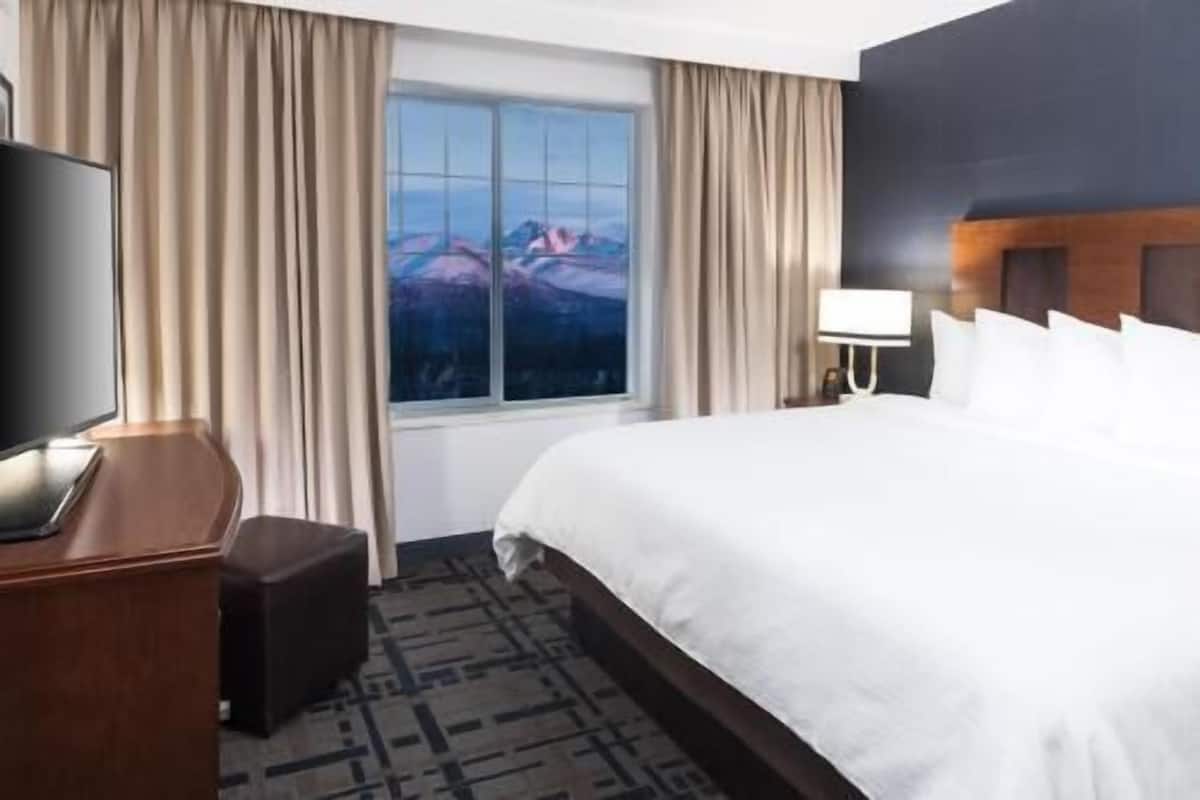 Quarto de hotel com cama, televisão, janela com cortinas e vista para montanhas.