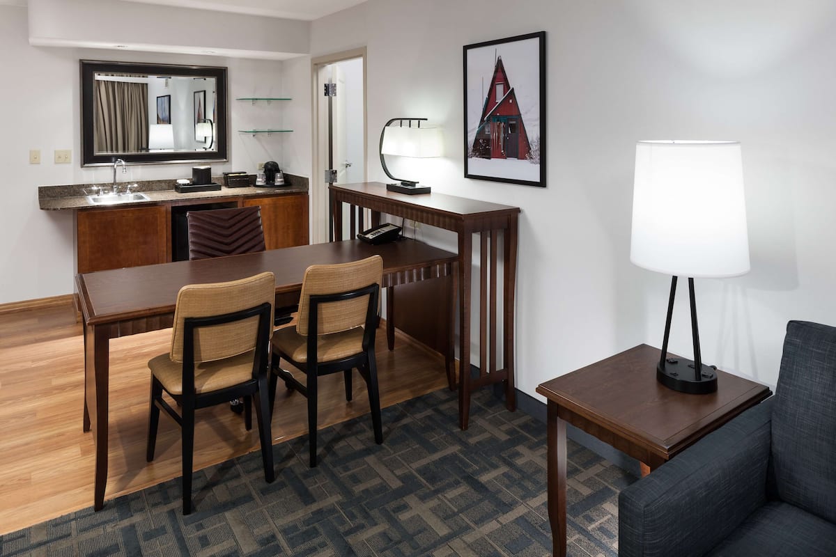 Quarto de hotel com área de jantar, uma mesa pequena com abajur, sofá e quadro na parede.