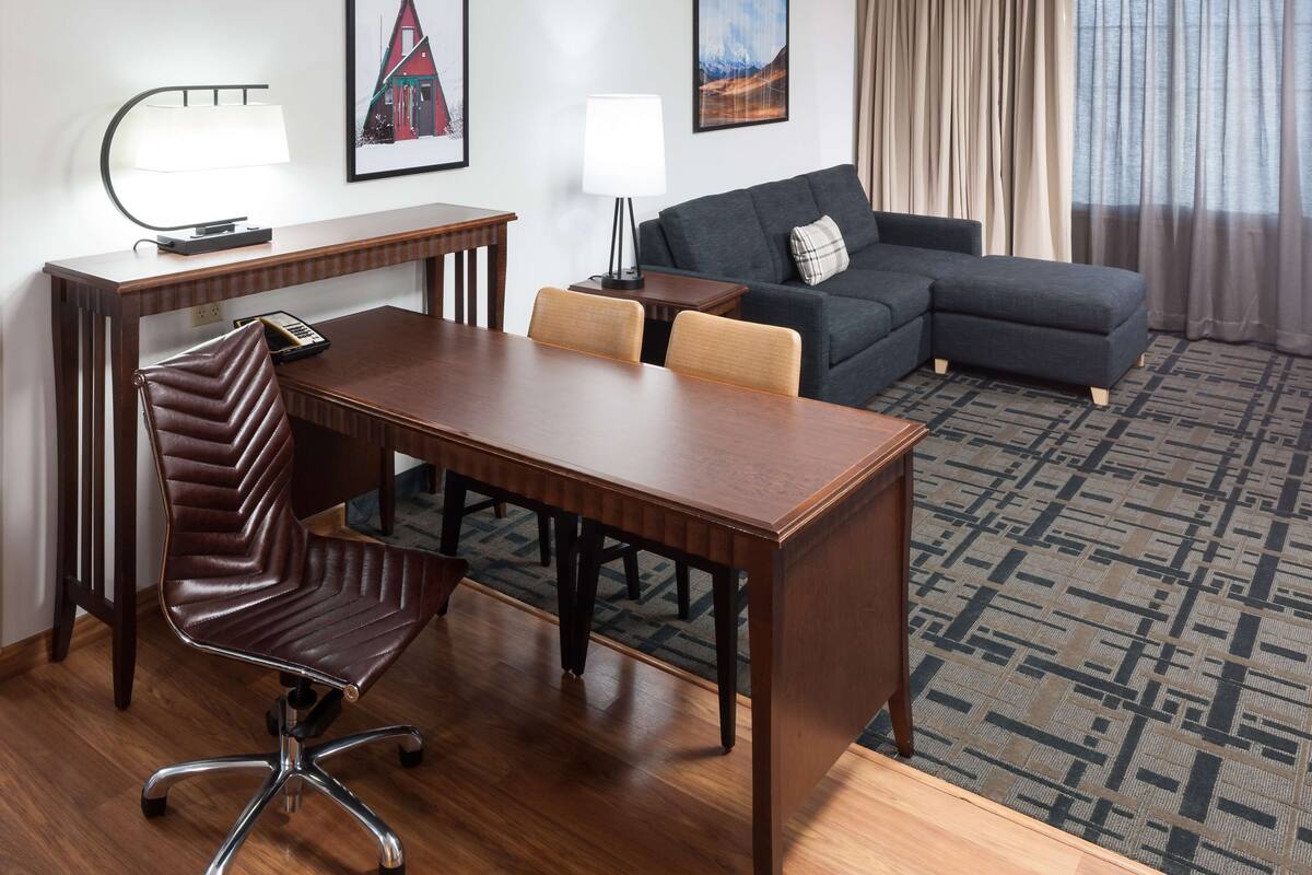Quarto de hotel com escrivaninha de madeira, cadeira de couro marrom, sofá cinza escuro, duas mesinhas de apoio e tapete estampado.