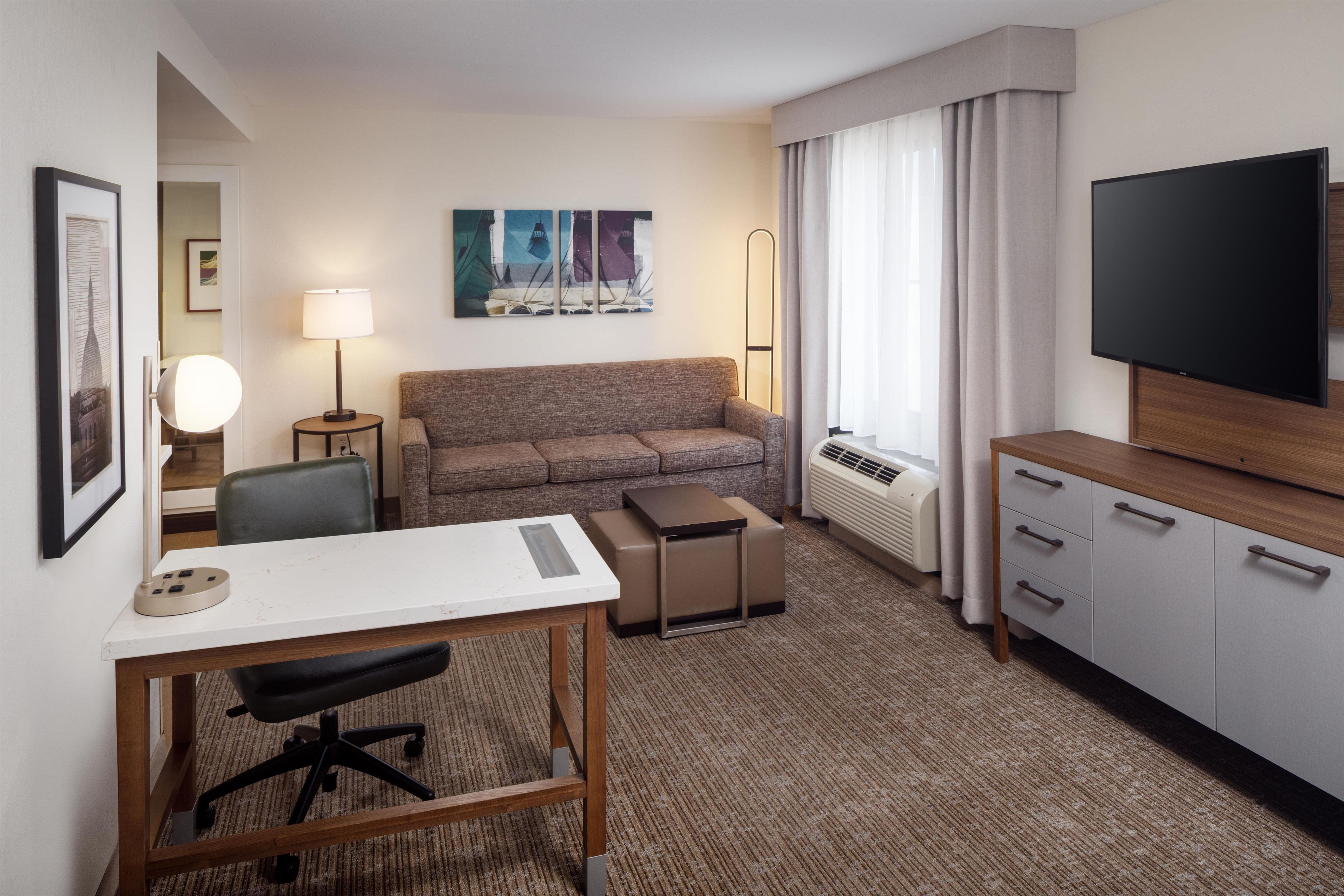 Quarto de hotel com escrivaninha, sofá, televisão e um quadro na parede.