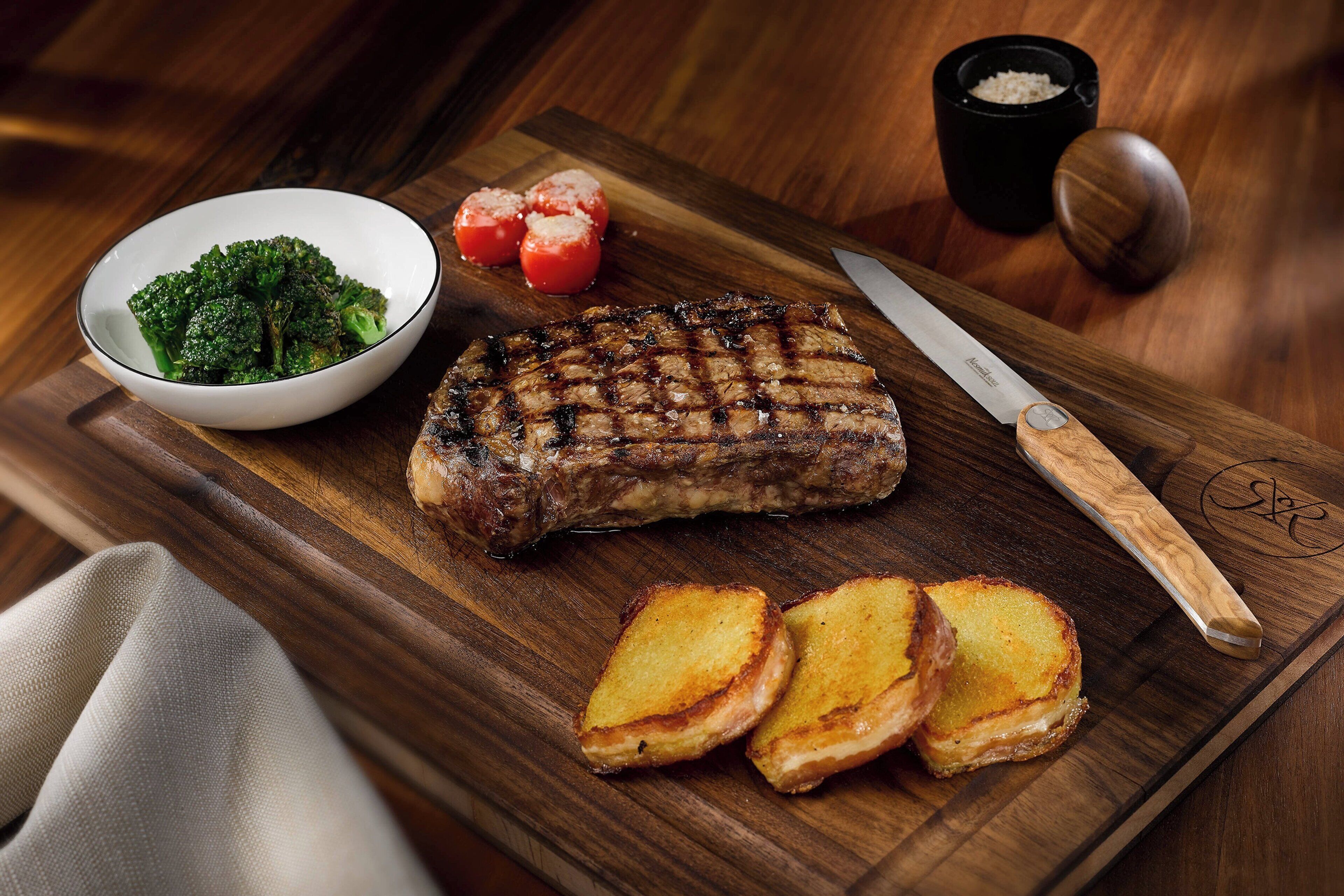 Tábua de madeira com bife grelhado, batatas assadas e uma tigela de brócolis.