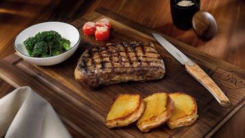 Tábua de madeira com bife grelhado, batatas assadas e uma tigela de brócolis.