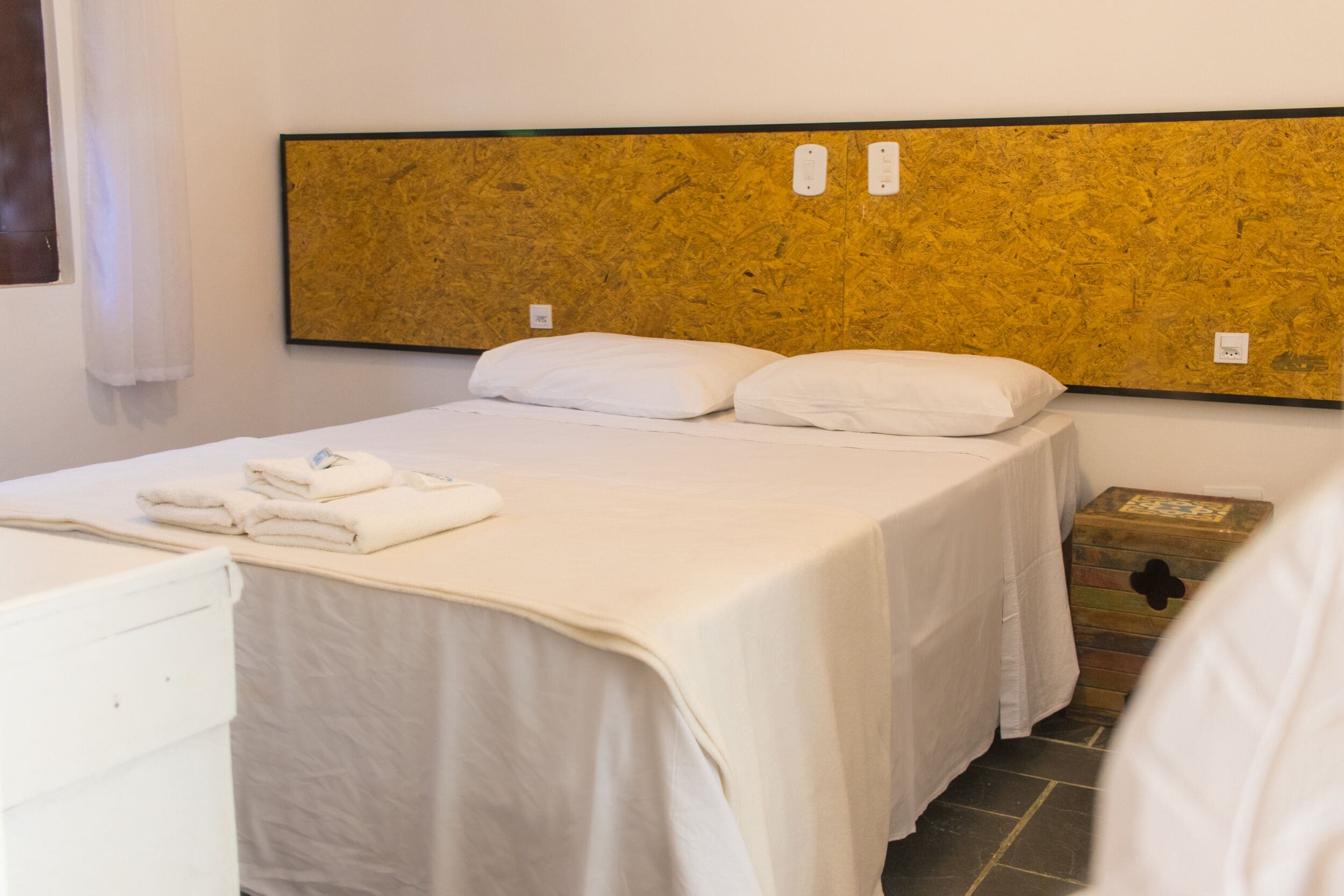 Uma cama de solteiro com lençóis brancos, cabeceira de madeira e um pequeno criado-mudo de madeira.