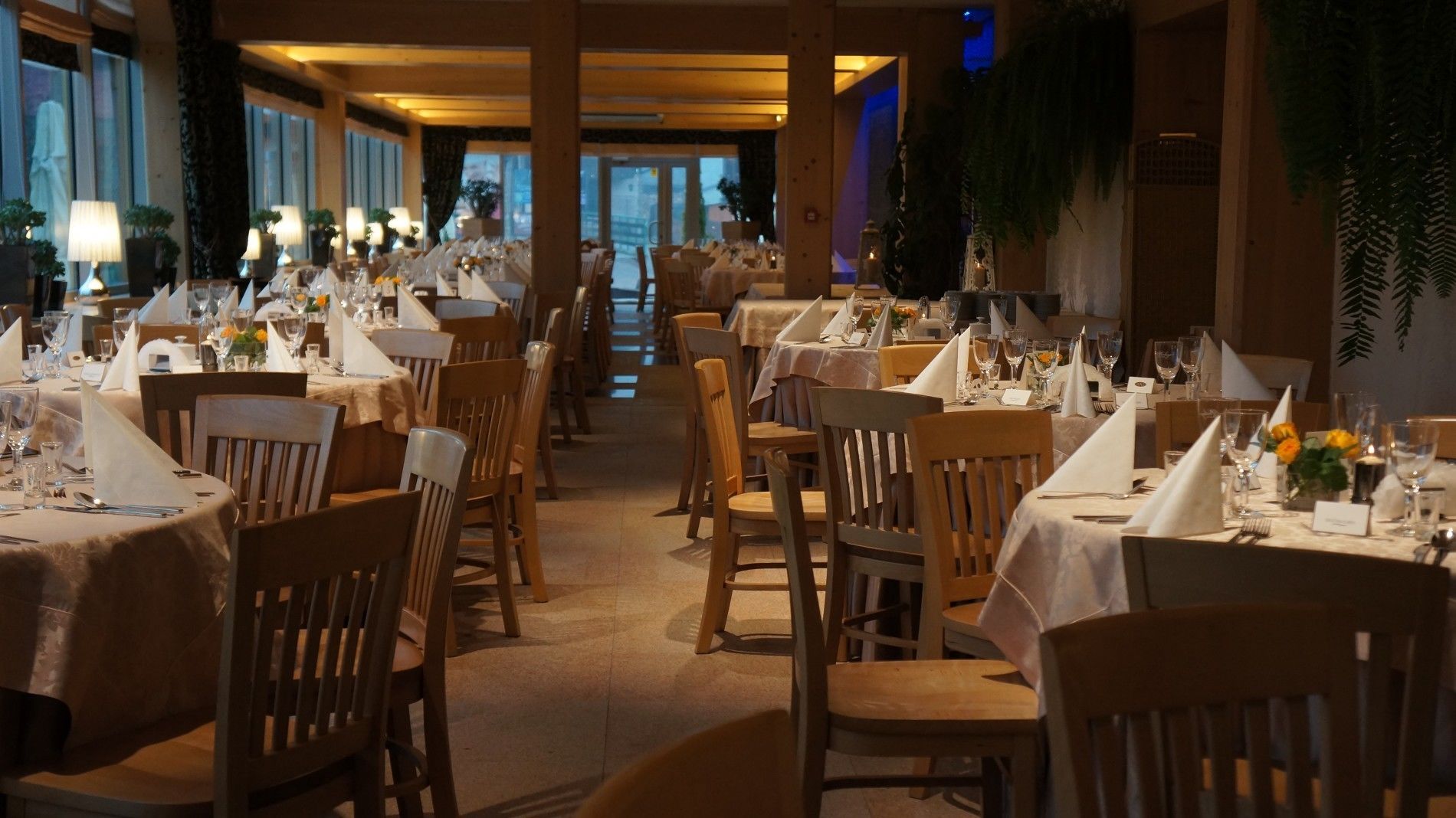 Restaurante com várias mesas redondas dispostas com toalhas brancas, cadeiras de madeira e copos.