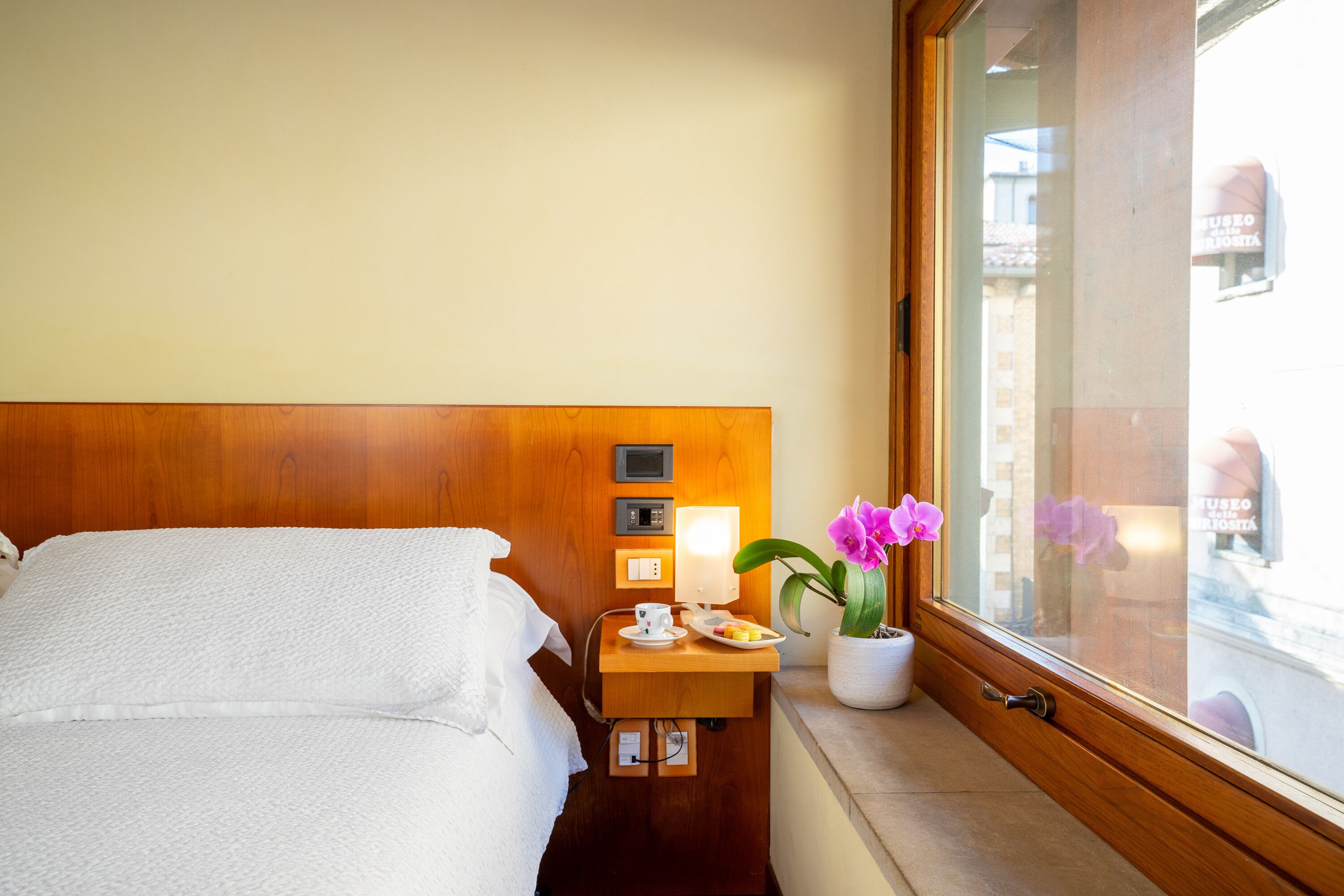 Quarto de hotel com cabeceira de madeira, mesa de cabeceira com abajur, uma orquídea em vaso pequeno e janela com vista para a rua.