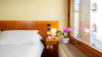 Quarto de hotel com cabeceira de madeira, mesa de cabeceira com abajur, uma orquídea em vaso pequeno e janela com vista para a rua.