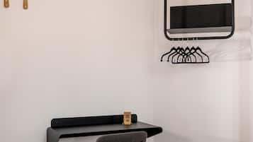 Quarto de hotel minimalista com uma cama, uma escrivaninha com cadeira, televisão na parede e um cabideiro.