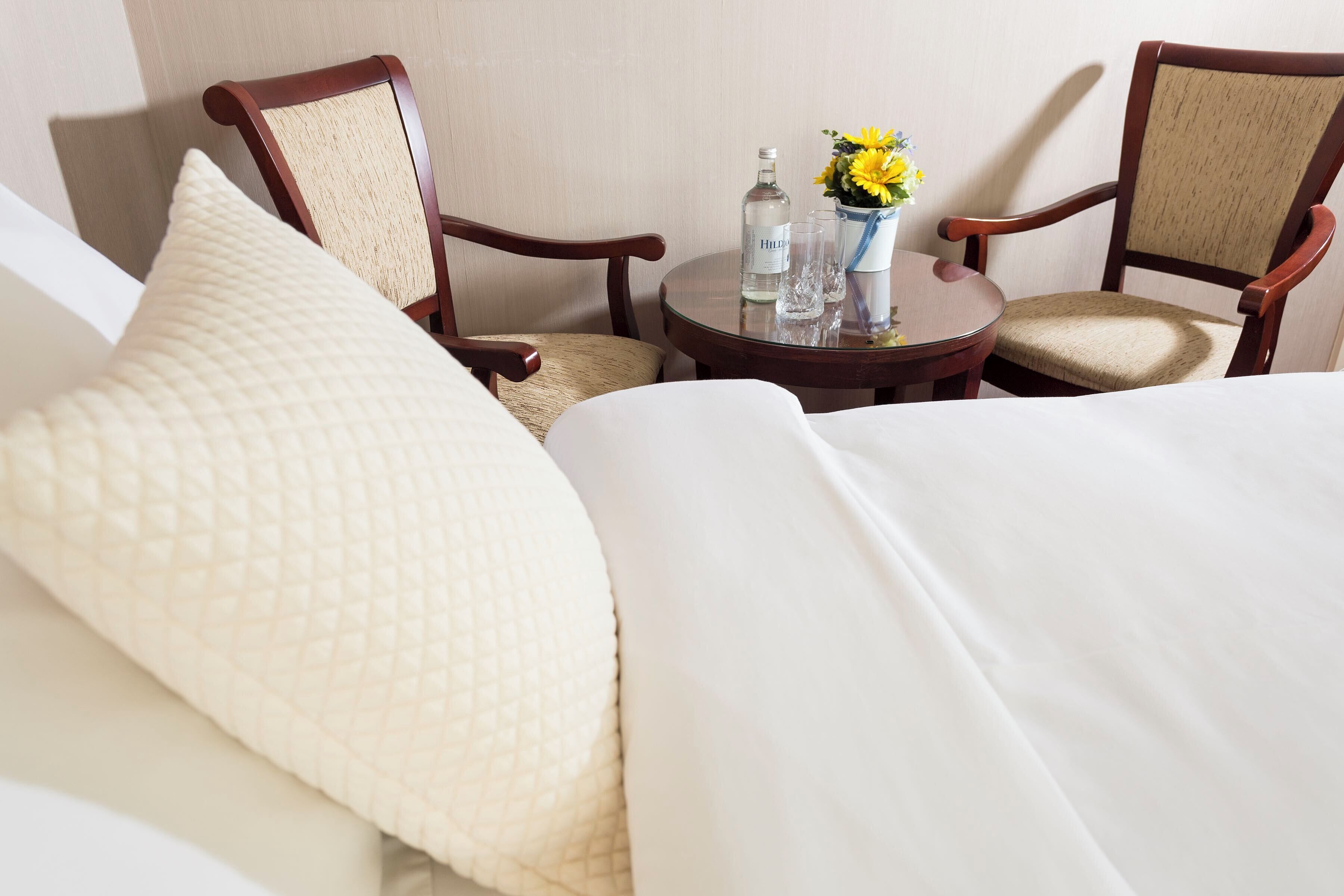 Quarto de hotel com uma cama, duas cadeiras de madeira, uma mesa redonda pequena com uma garrafa, copos e um vaso de flores.