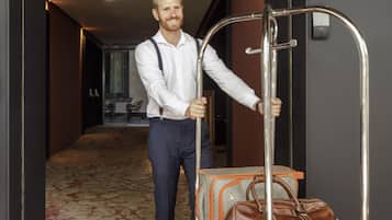 Um homem de camisa branca e calças azuis empurra um carrinho de bagagem com uma mala e uma bolsa de couro marrom.