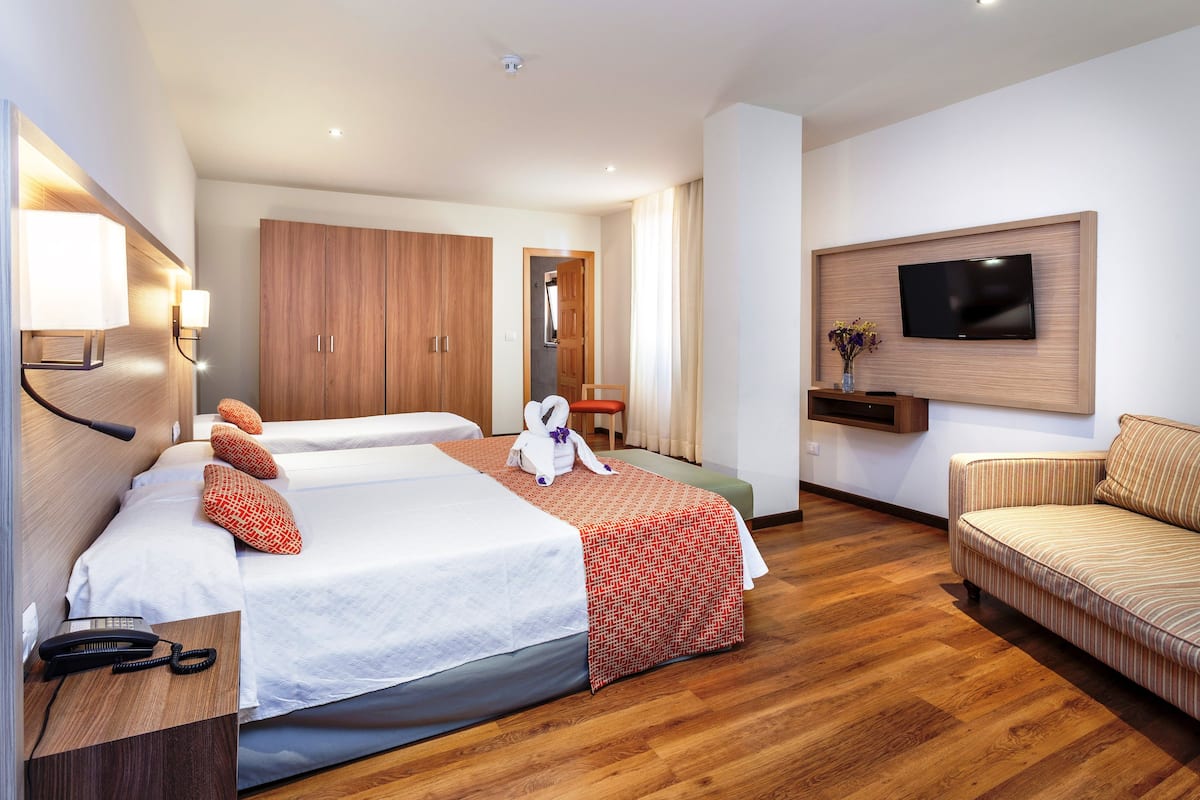 Quarto de hotel com duas camas, um sofá, uma TV e um guarda-roupa de madeira.
