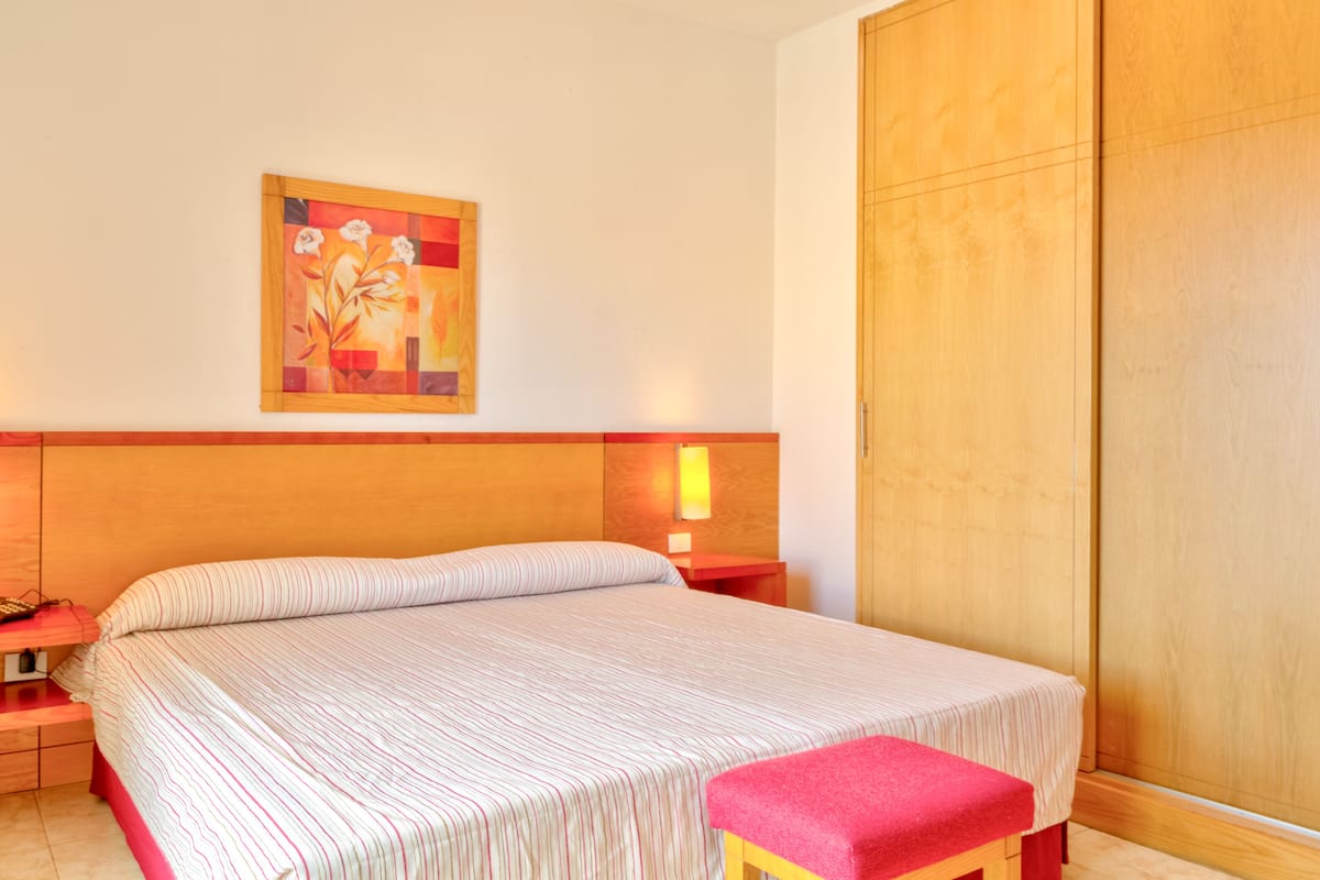 Cama bem arrumada com colcha listrada, mesa de cabeceira com abajur e guarda-roupa de madeira.