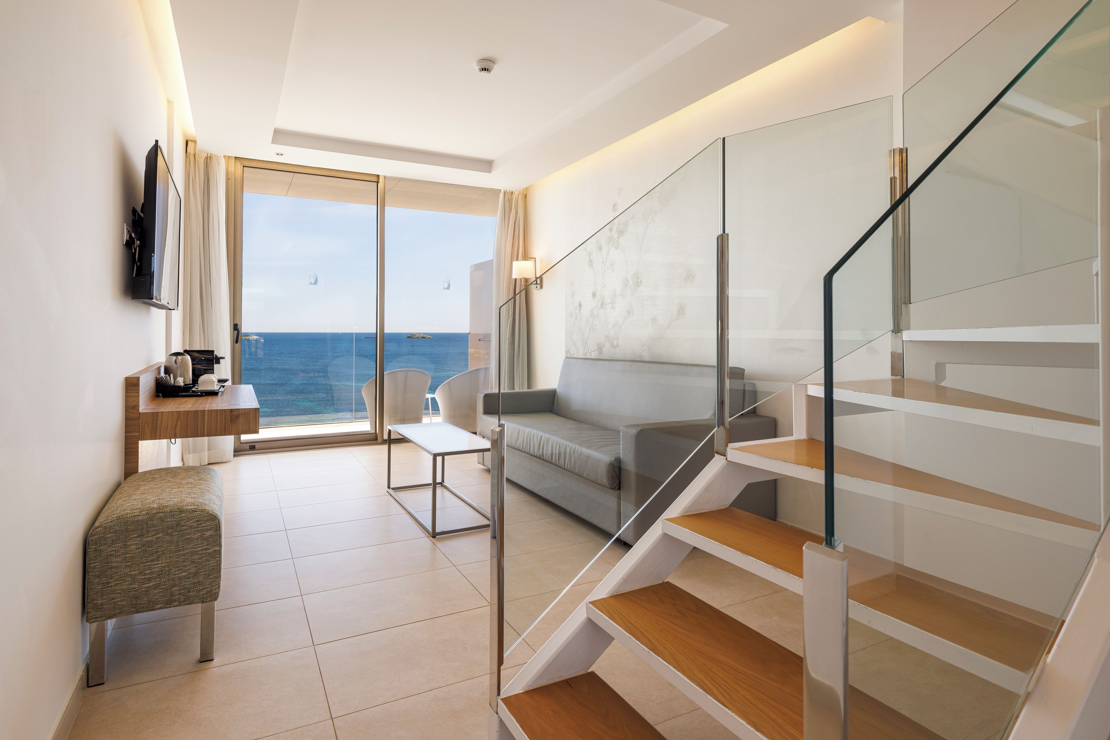 Quarto de hotel moderno com escada de vidro, um sofá, uma mesa pequena e um banco.