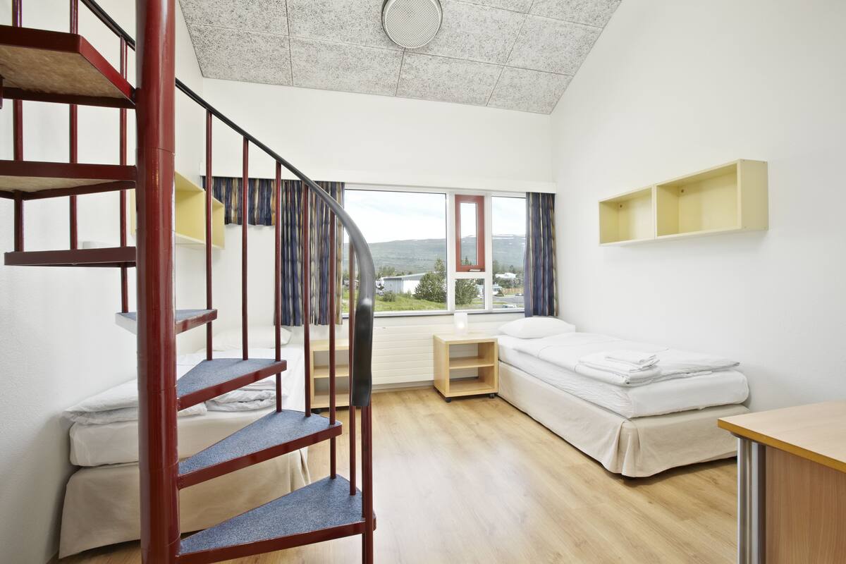 Quarto moderno com escada de madeira, uma cama, uma escrivaninha e janelas amplas com vista para o exterior.