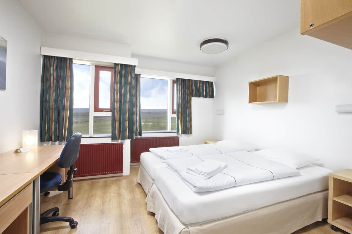 Quarto de hotel com cama, escrivaninha, cadeira, janela com cortinas, radiador e prateleira.