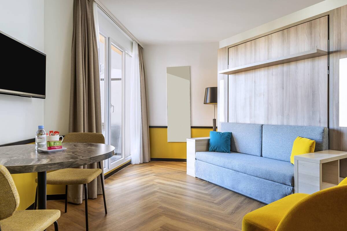 Quarto de hotel moderno com área de jantar, sofá com almofadas, televisão de tela plana e uma grande janela com cortinas.
