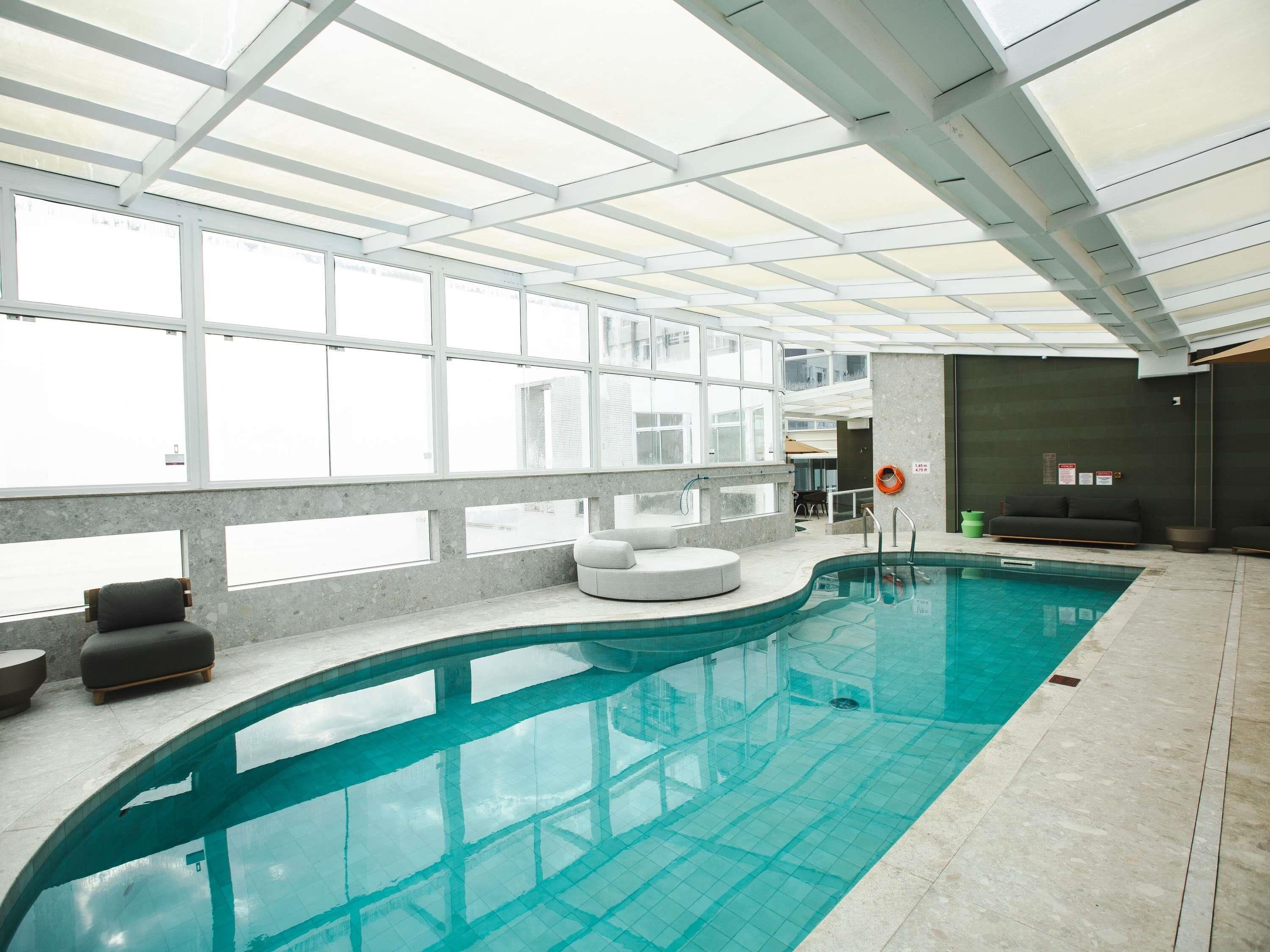Piscina interna com teto de vidro, área de estar com assentos e vista para a paisagem urbana.
