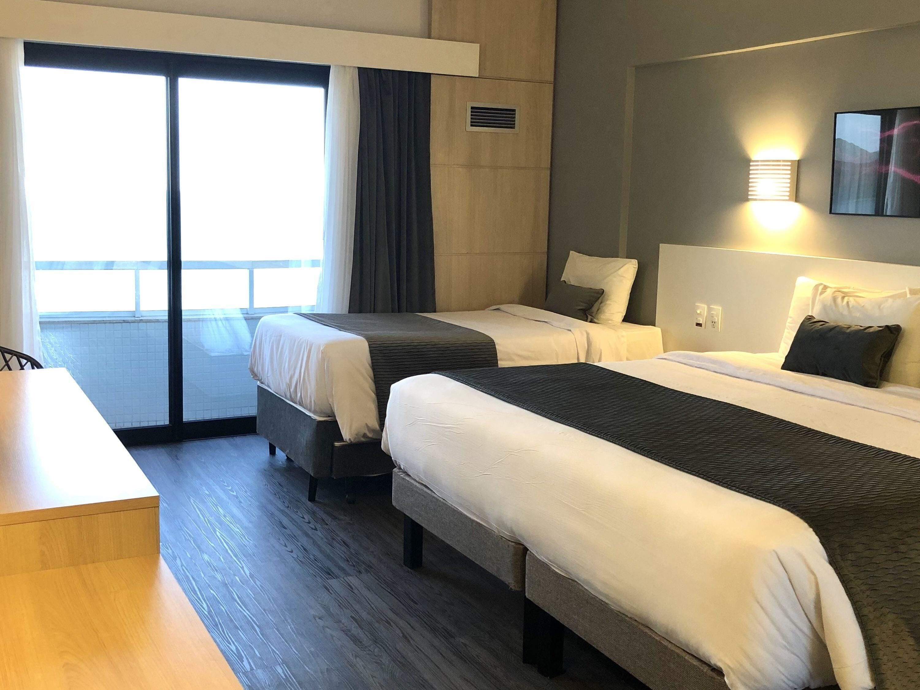Quarto de hotel com duas camas, uma escrivaninha e uma janela grande com cortinas.