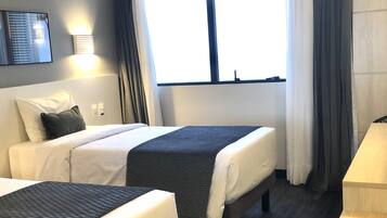 Quarto de hotel com duas camas, uma janela grande com cortinas, uma luminária fixada na parede e uma escrivaninha de madeira.